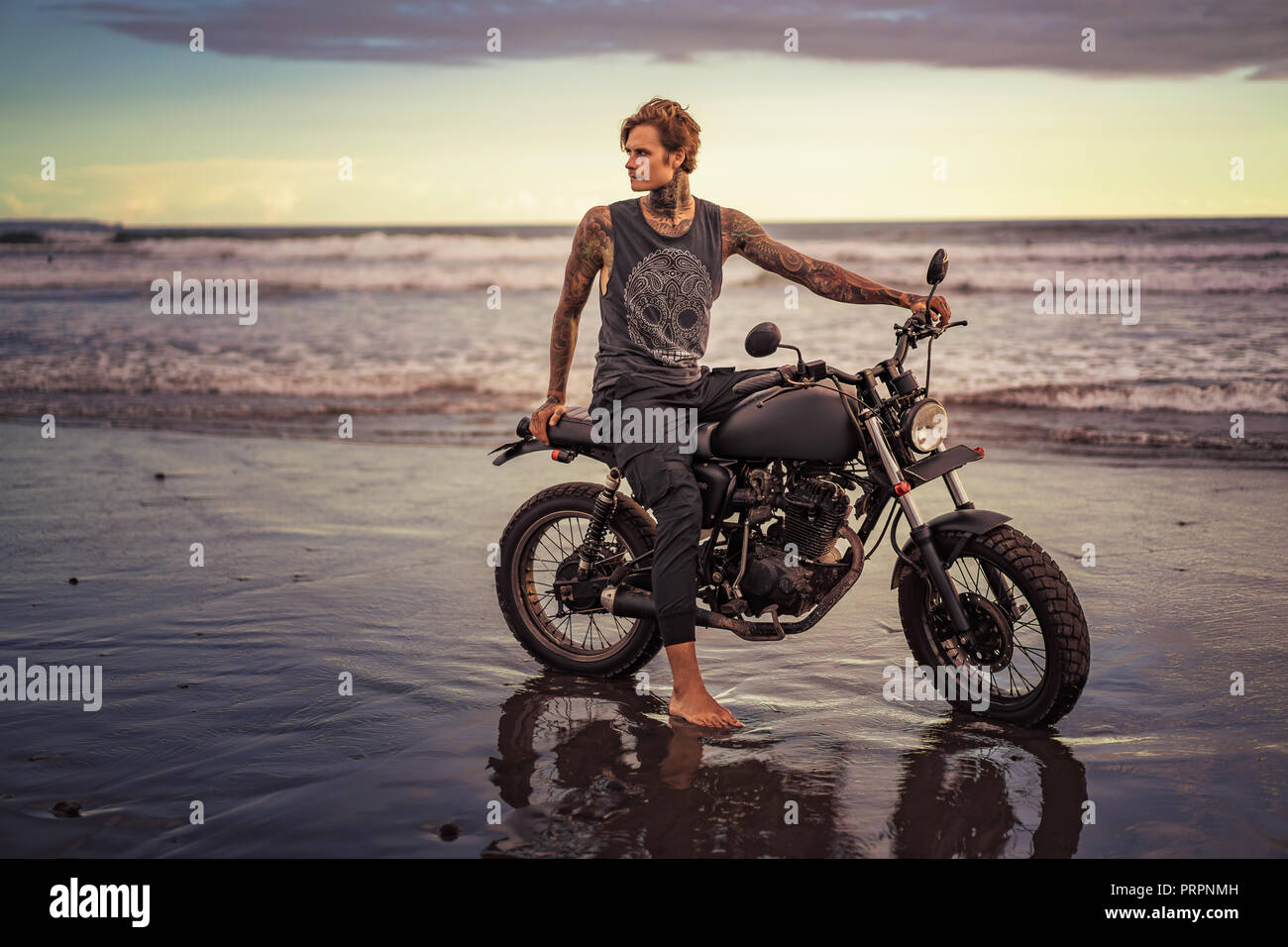tattooed biker sitting on motorcycle on ocean beach and looking away ...