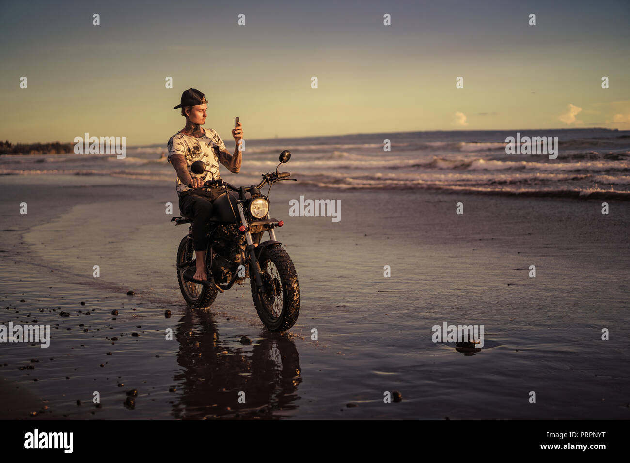 tattooed biker sitting on motorbike on ocean beach and taking photo of ...