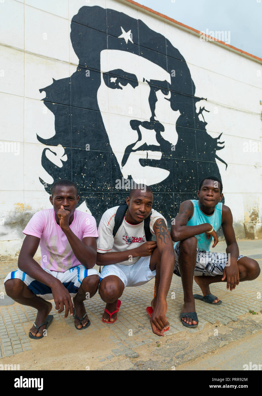 Three angolan men squatting in front of a wall painting of Che Guevara ...