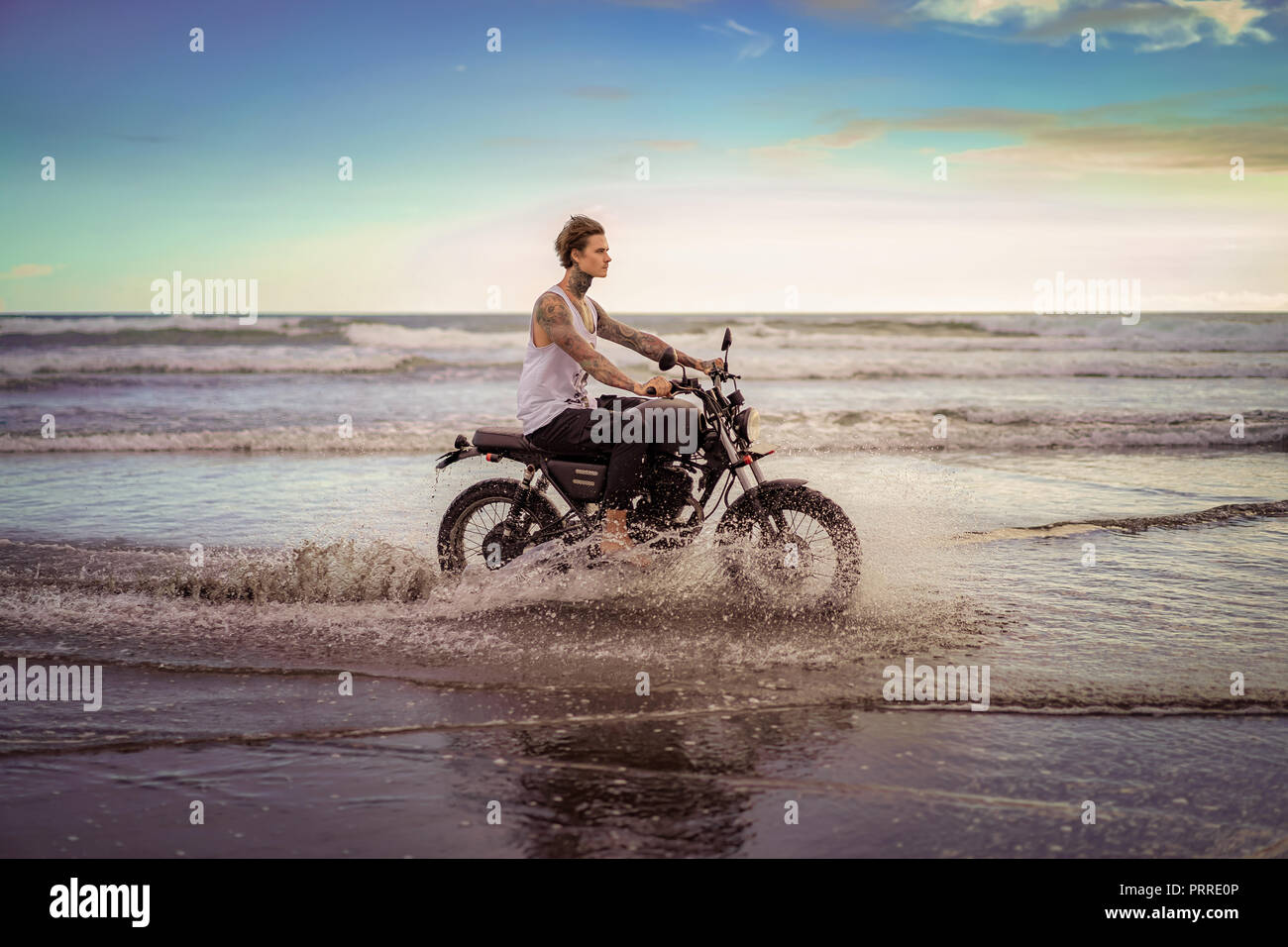 side view of handsome tattooed man riding motorcycle in ocean waves on ...