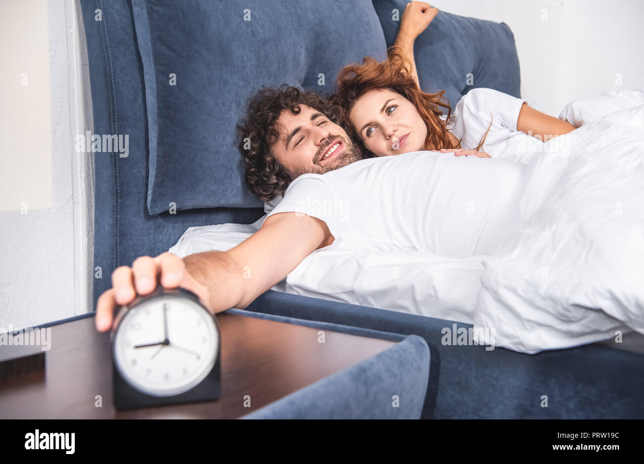 happy young couple waking up together, man reaching to alarm clock ...