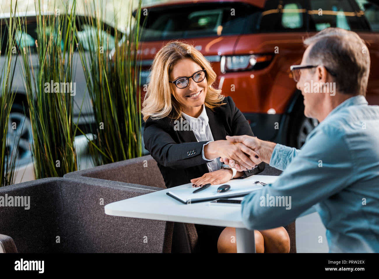 happy adult customer and female car dealer shaking hands in showroom ...