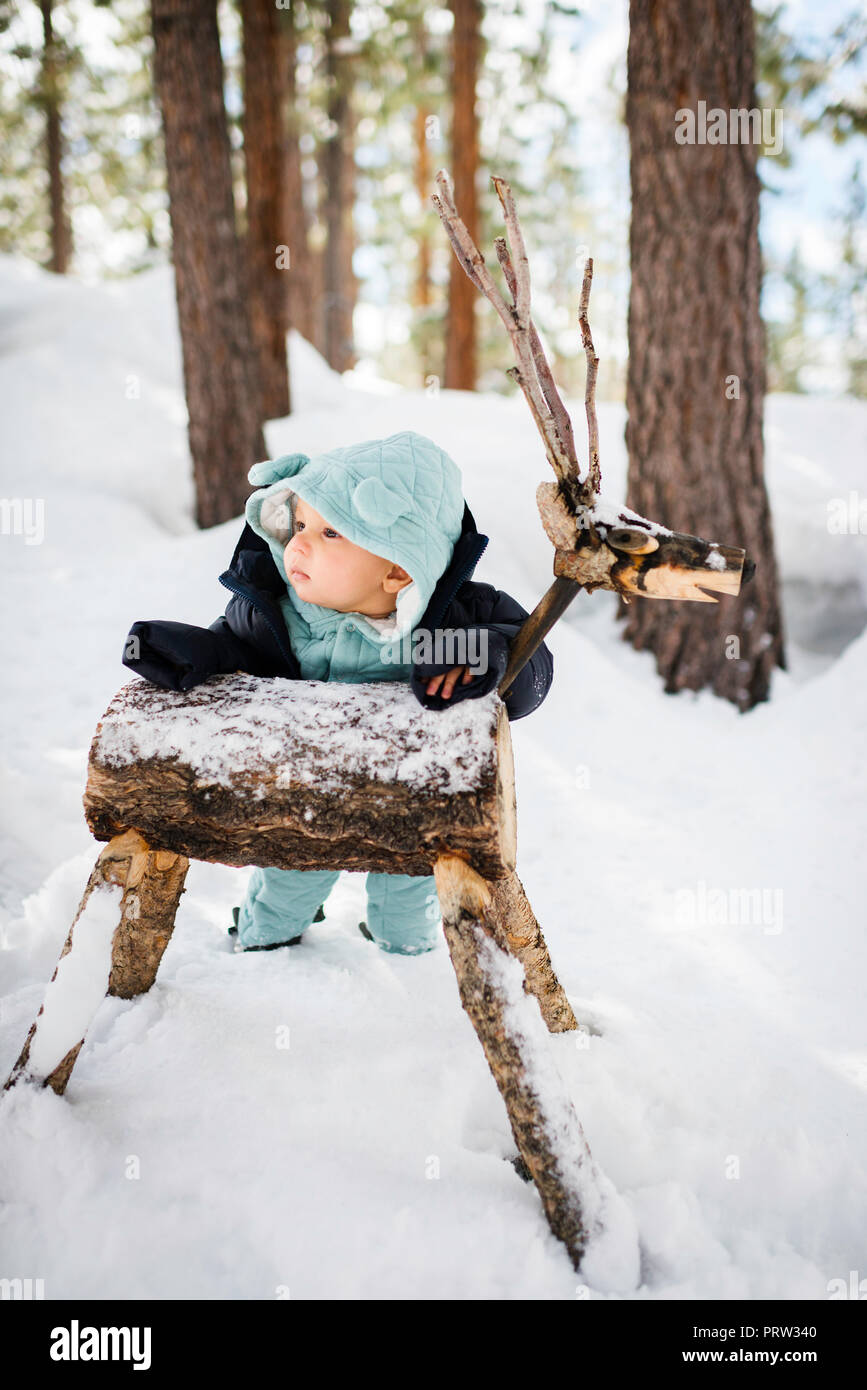 Baby boy ankle deep in forest snow leaning against wooden deer, South ...