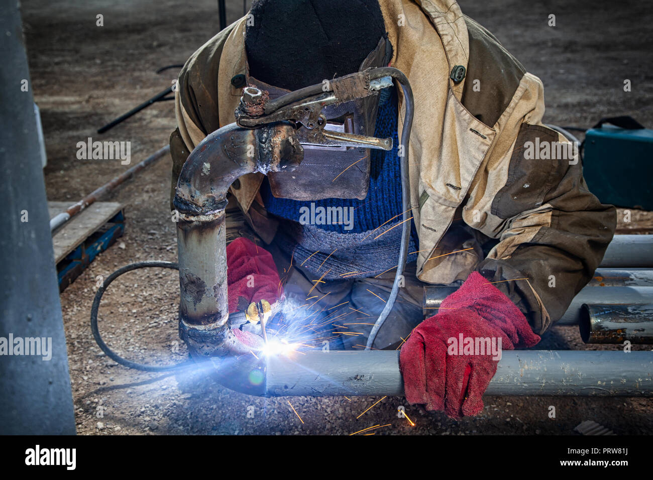 Close up welder weld metal pipe at construction site. A builder in brown uniform, welding mask ...