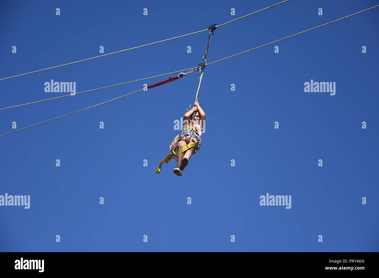 Happily smiling little girl hanging on a zip-line high in the blue sky ...