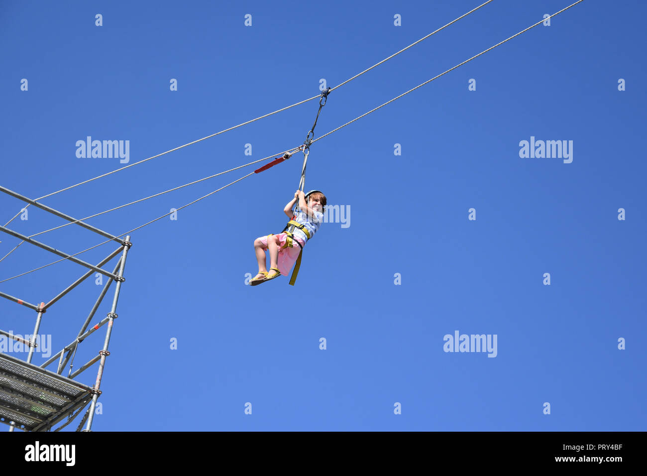 Happily smiling little girl hanging on a zip-line high in the blue sky ...
