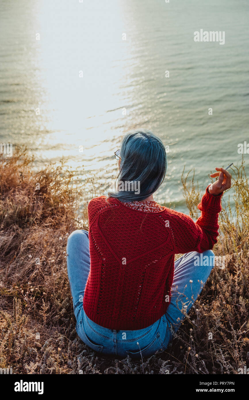 A young rebel girl in a red sweater smoking on a rocky coast near the ...