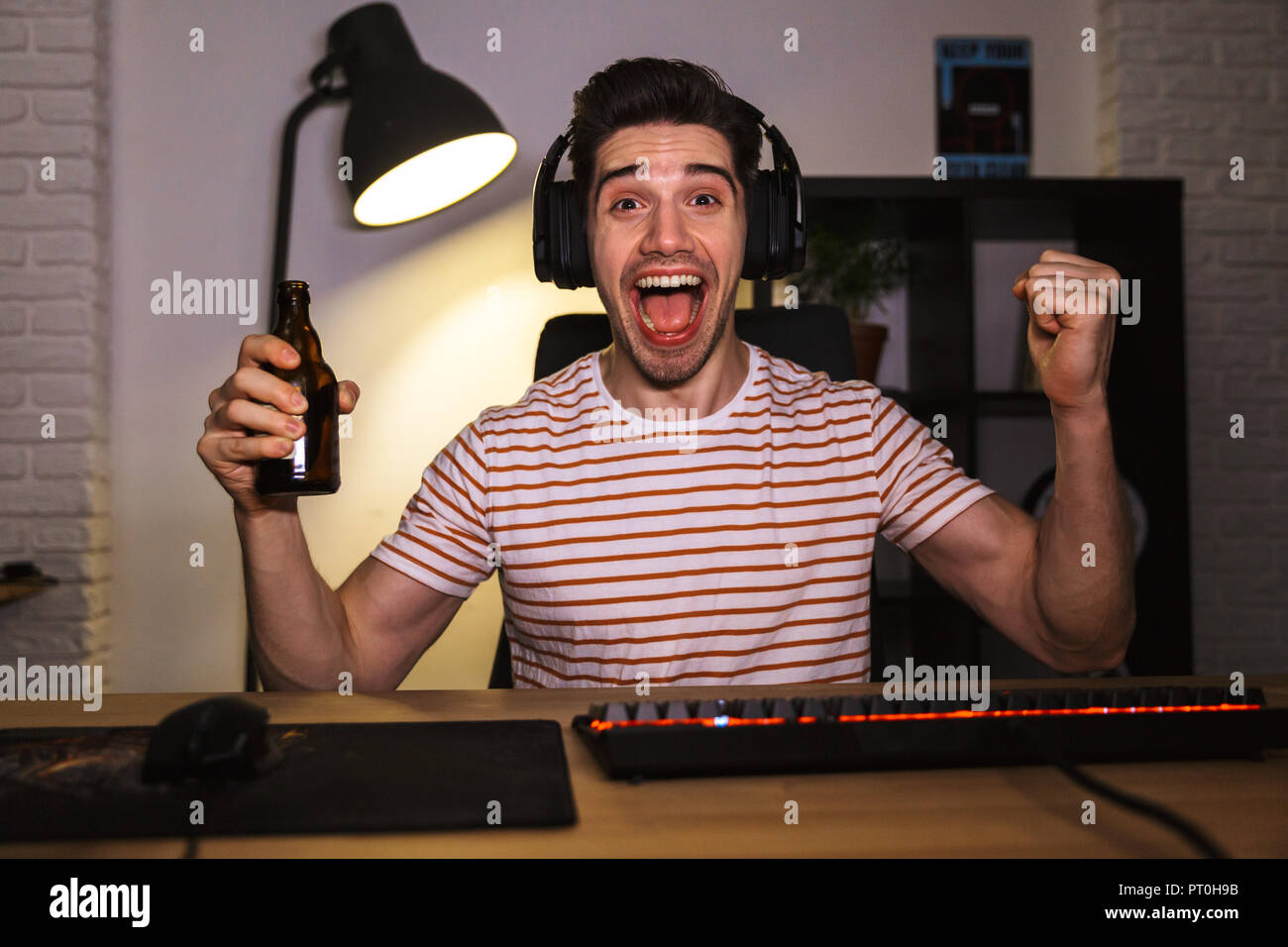 Portrait of joyful gamer guy wearing headphones screaming and drinking ...