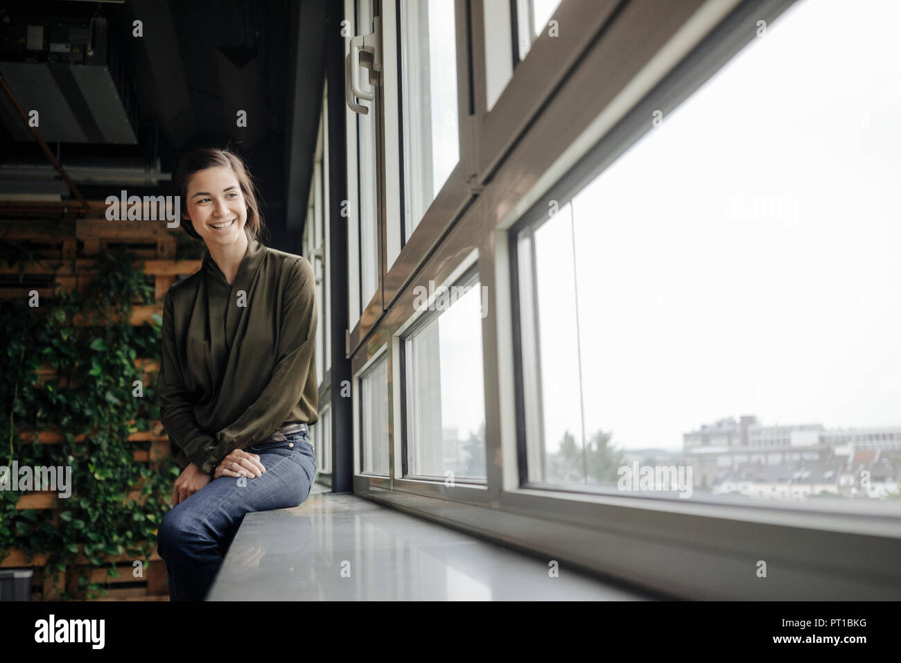 Happy young woman in office looking out of window Stock Photo - Alamy