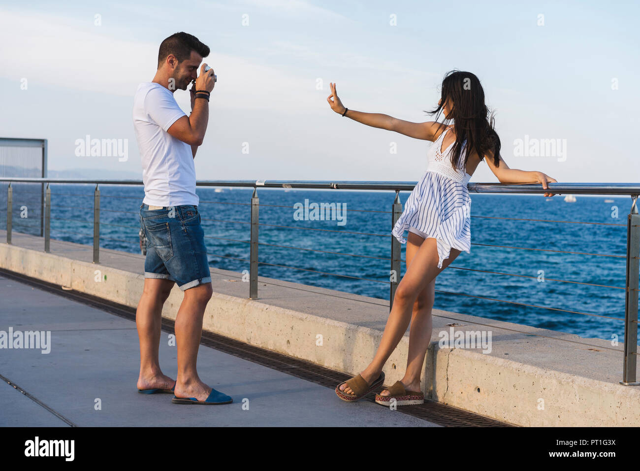 Young urban couple taking pictures by the sea, woman making peace sign ...