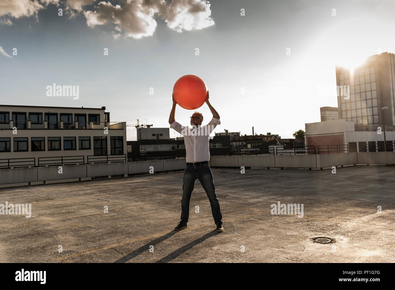 Mature man playing with orange fitness ball on rooftop of a high-rise ...