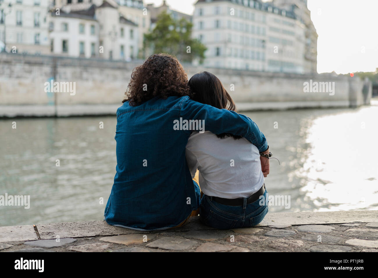 France, Paris, rear view of young couple sitting on a wall at river ...