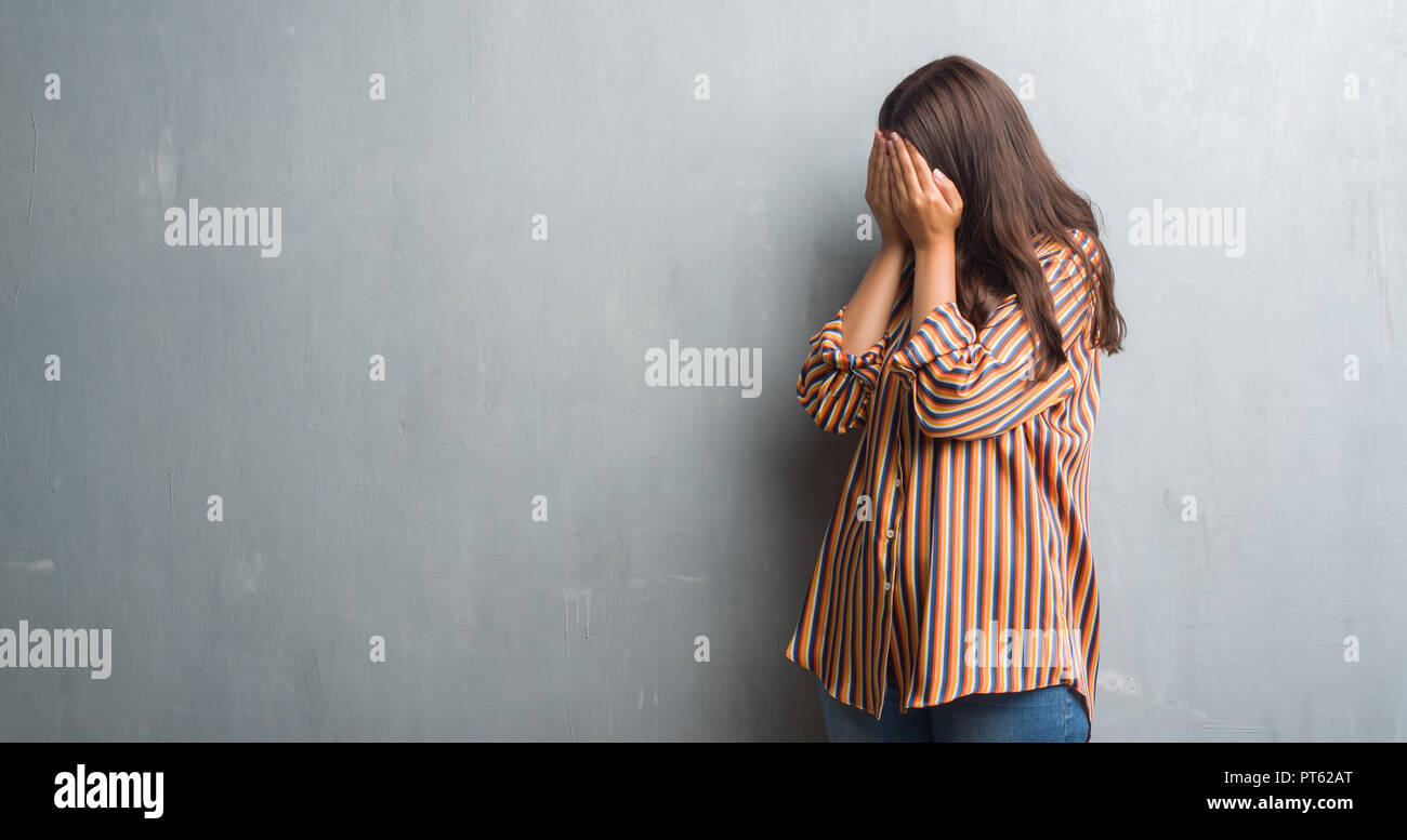 Young brunette woman over grunge grey wall with sad expression covering ...