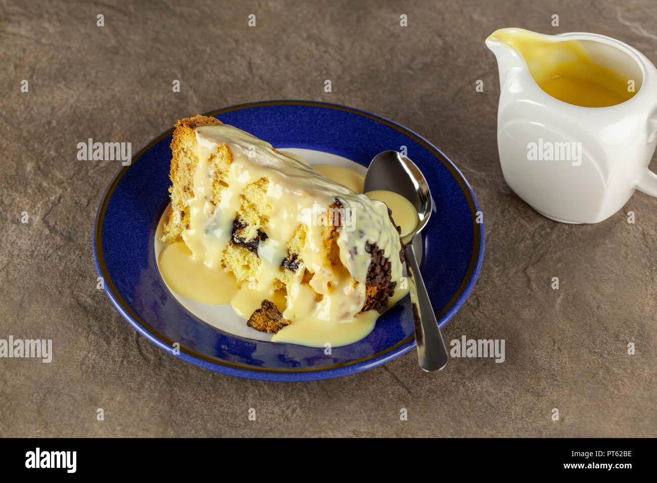 Chocolate topped sponge cake and custard on a blue plate with a jug of ...