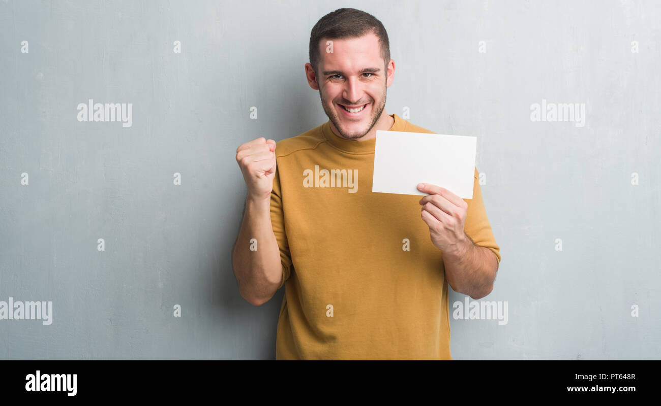 Young caucasian man over grey grunge wall holding blank card screaming ...
