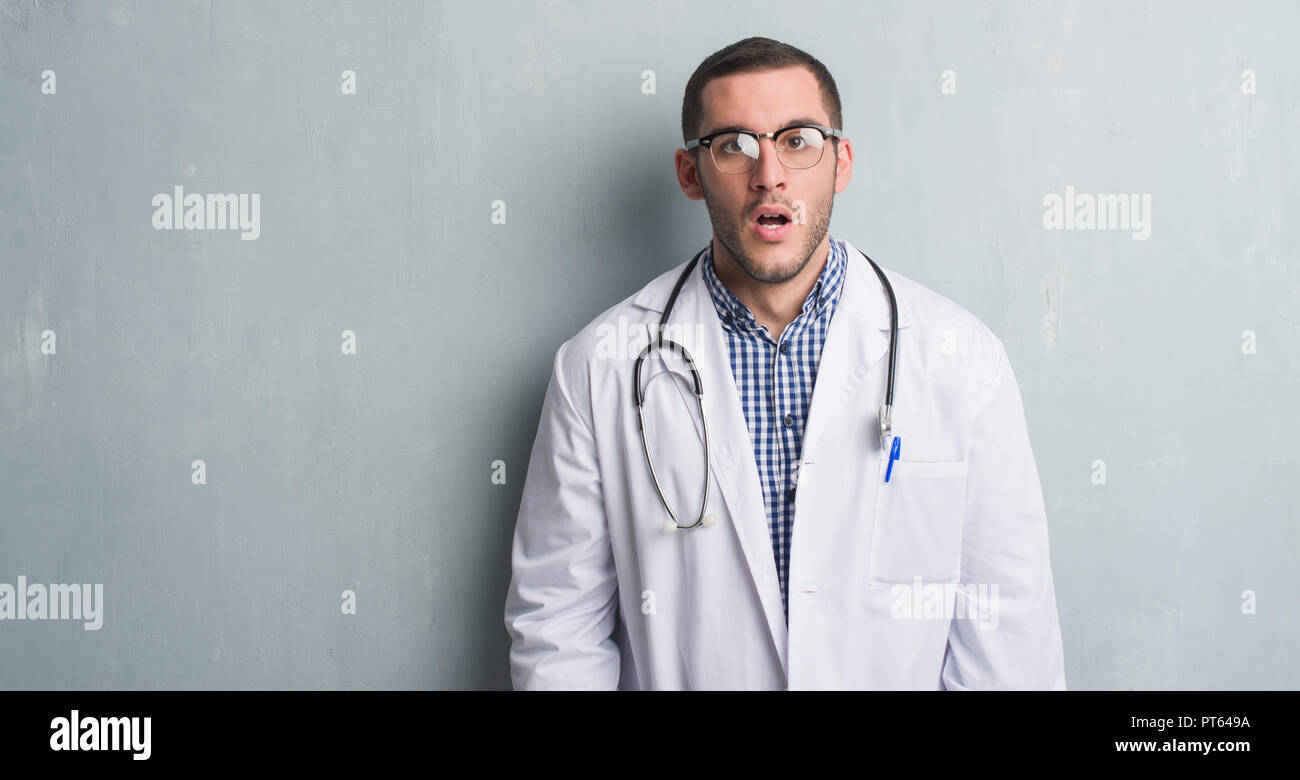 Young caucasian man over grey grunge wall wearing doctor uniform scared ...