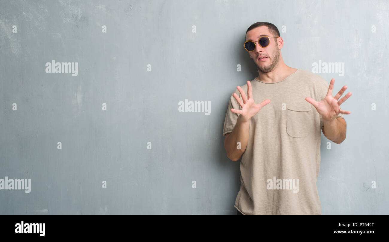 Young caucasian man over grey grunge wall wearing sunglasses afraid and ...
