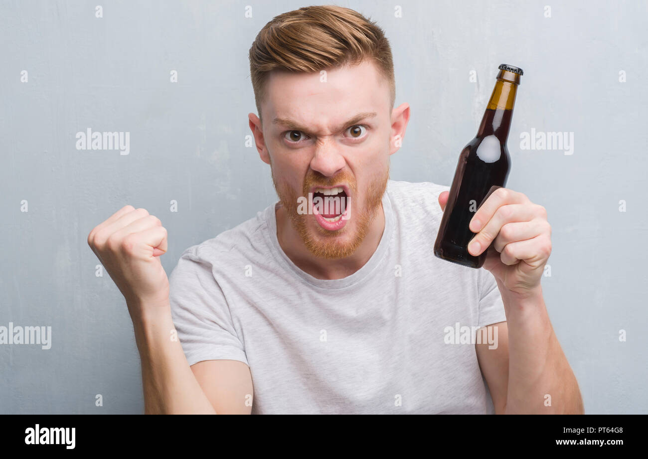 Young redhead man over grey grunge wall drinking beer bottle annoyed ...