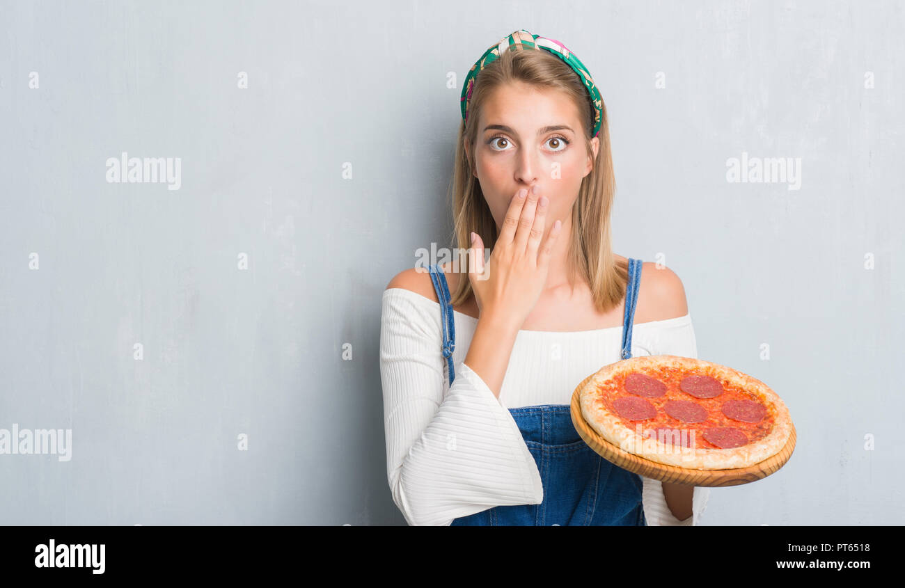Beautiful young woman over grunge grey wall eating pepperoni pizza ...