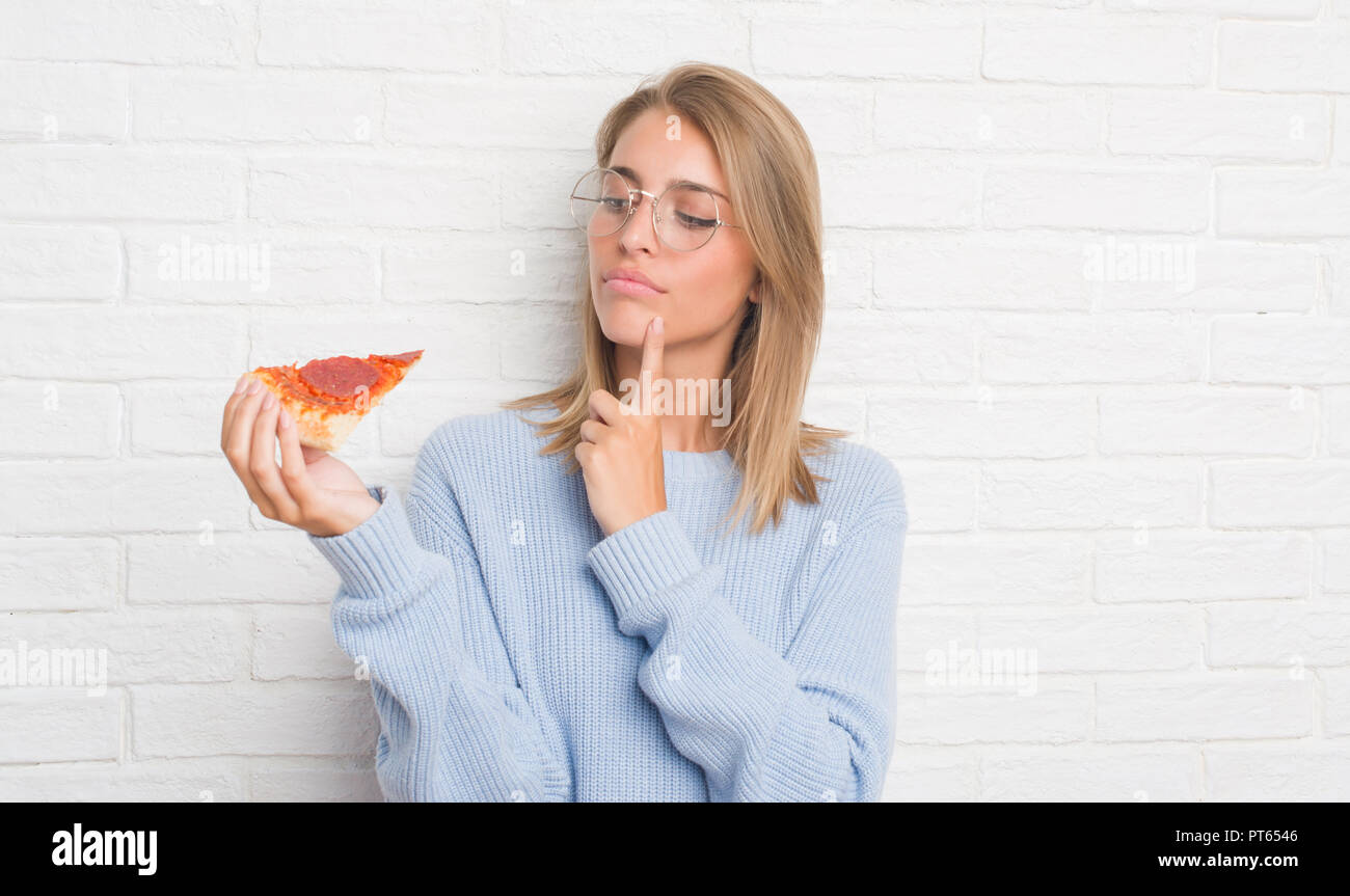 Beautiful young woman over white brick wall eating pizza slice serious ...