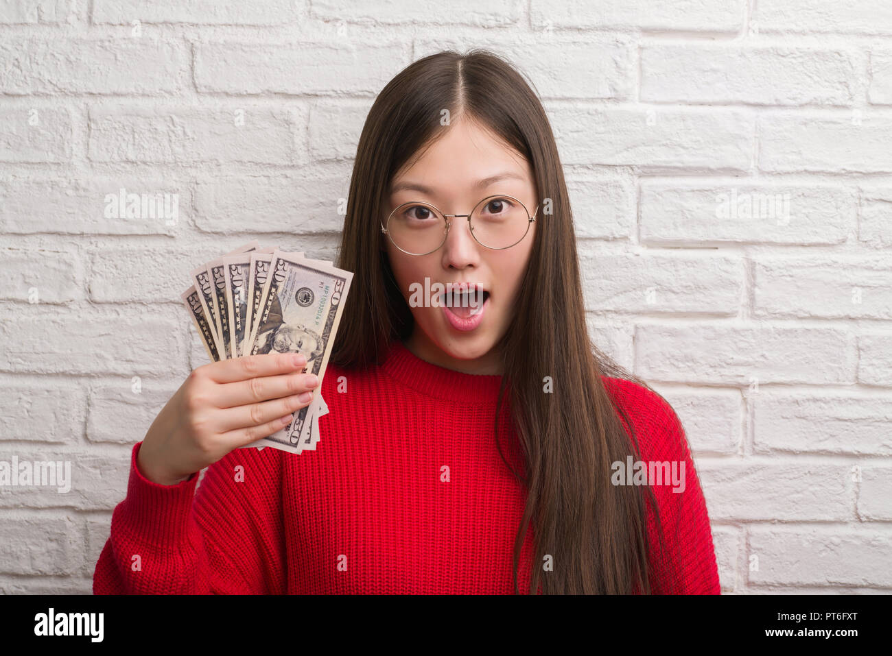 Young Chinese woman over brick wall holding dollars scared in shock ...