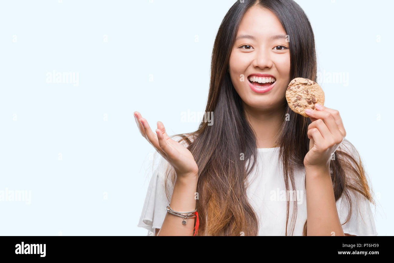 Young asian woman eating chocolate chip cookie over isolated background ...