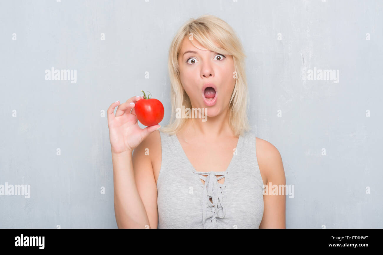 Adult caucasian woman over grunge grey wall holding fresh tomato scared ...