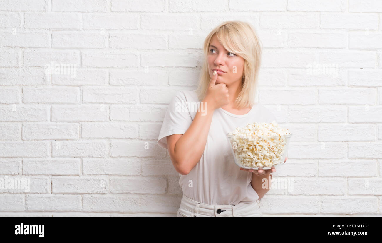 Adult caucasian woman over white brick wall eating pop corn serious ...