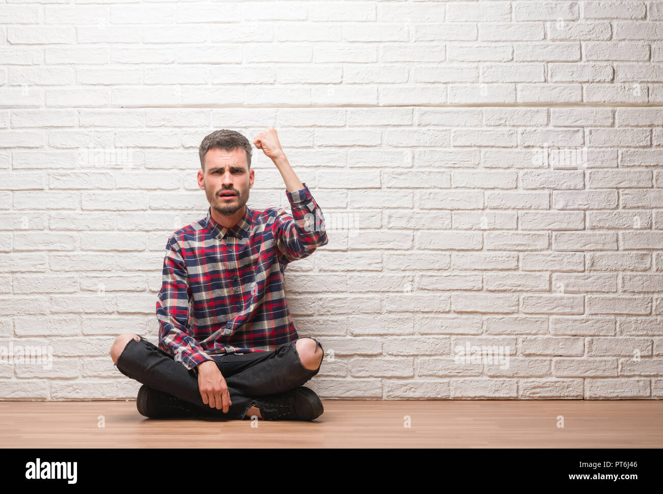 Young adult man sitting over white brick wall annoyed and frustrated ...