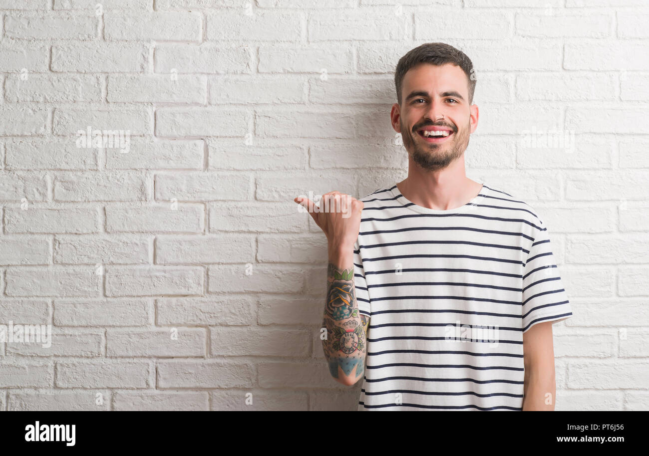 Young adult man standing over white brick wall pointing and showing ...