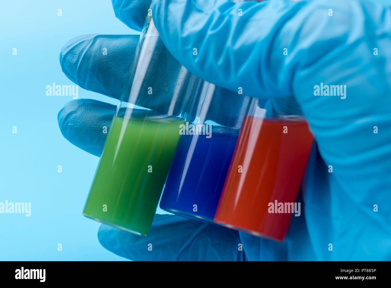 A gloved hand holds three vials of different liquid inside on a blue ...