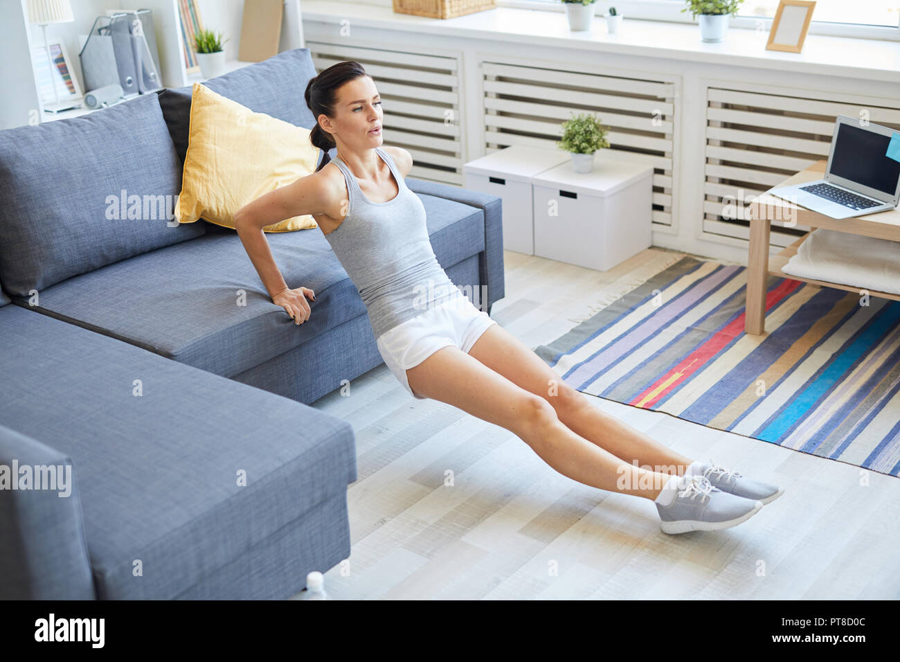 Fit young sportswoman doing physical exercises by sofa in living-room ...