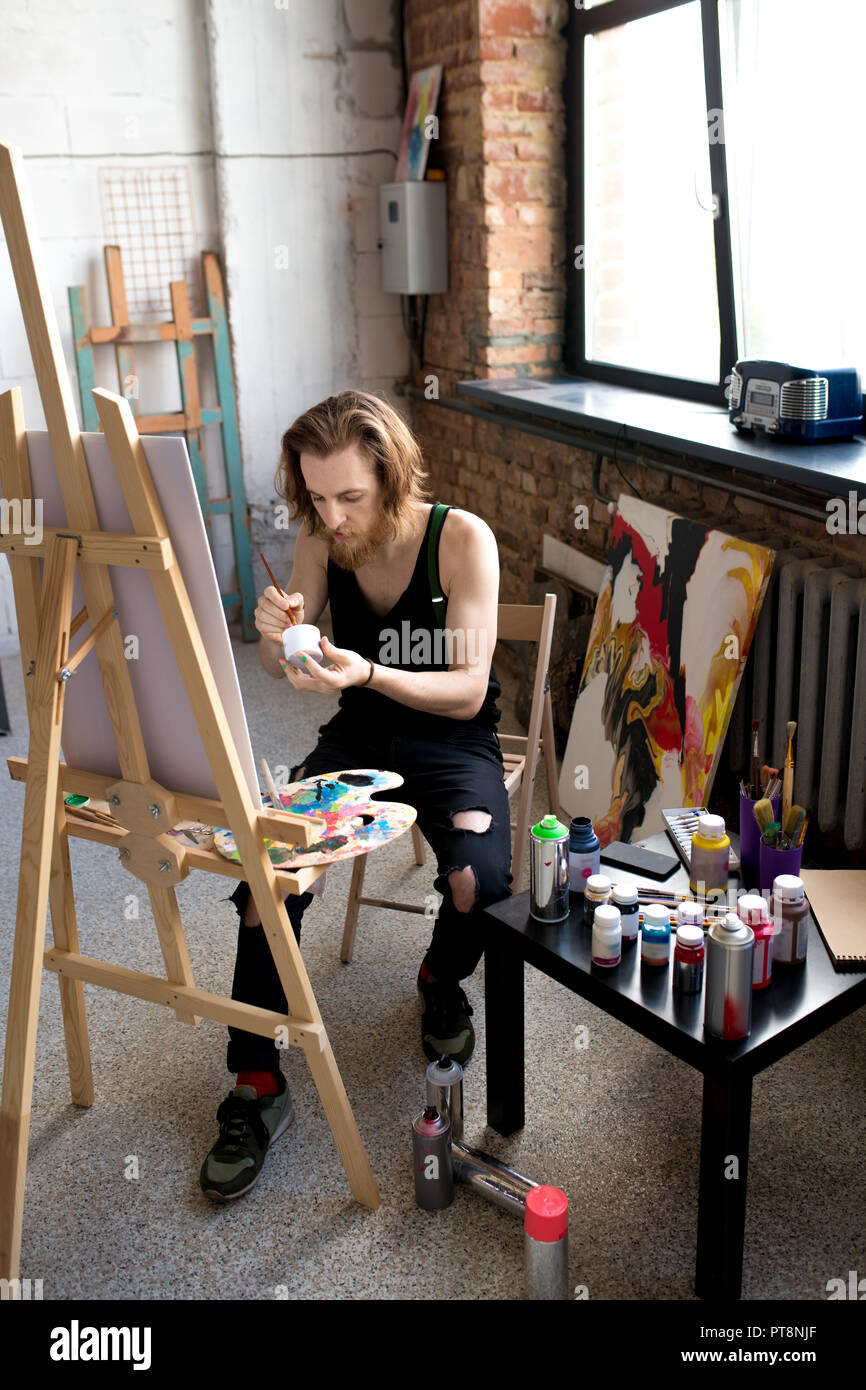 Full length portrait of contemporary long haired male artist painting ...