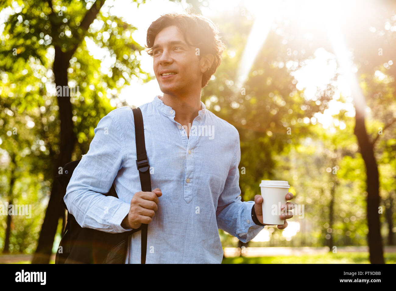 Smiling young casual man drinking coffee while walking at the city park ...
