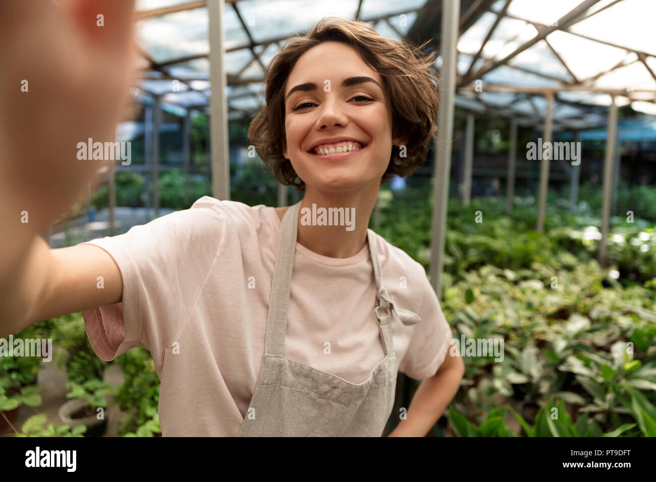 Image of beautiful cute woman gardener standing over plants in greenhouse take a selfie by ...