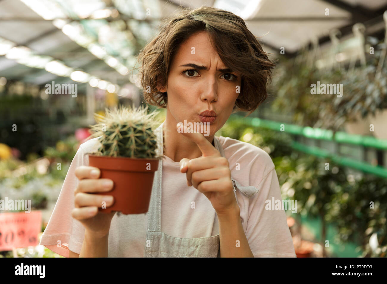 Image of scared emotional cute woman gardener standing over flowers plants in greenhouse holding ...
