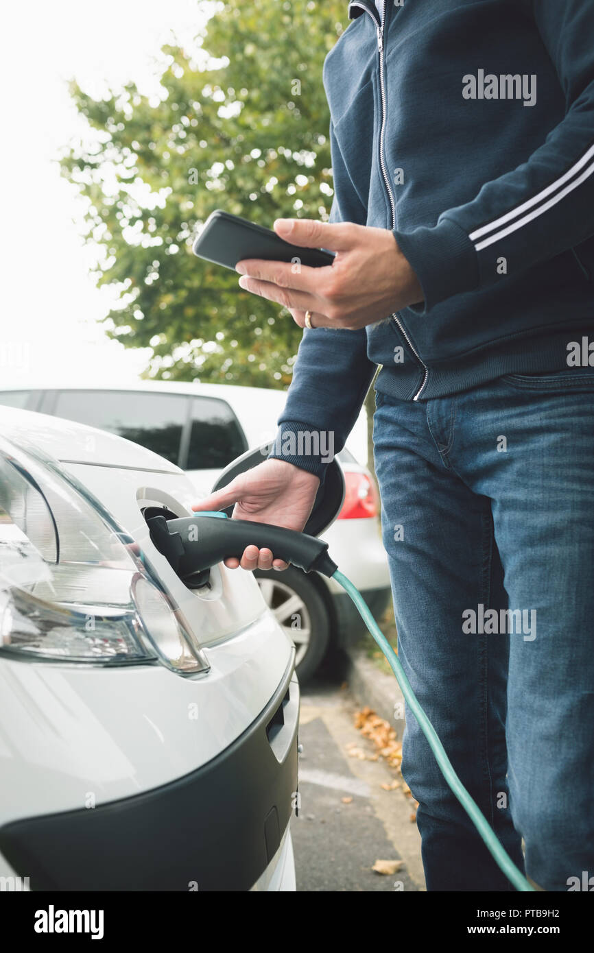 Man using mobile phone while charging electric car at charging station ...