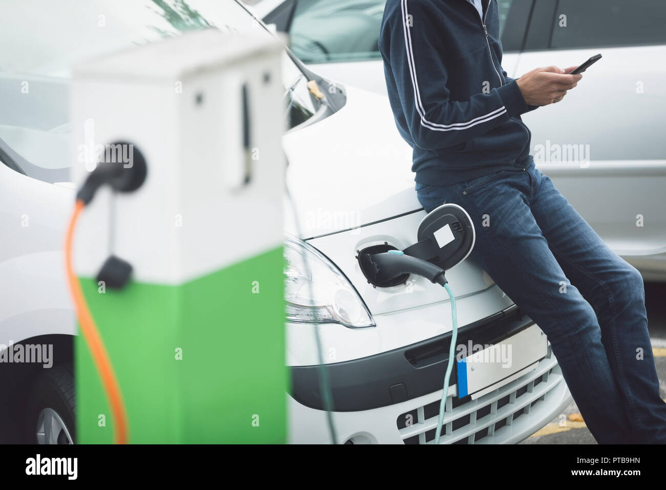 Man using mobile phone while charging electric car at charging station ...