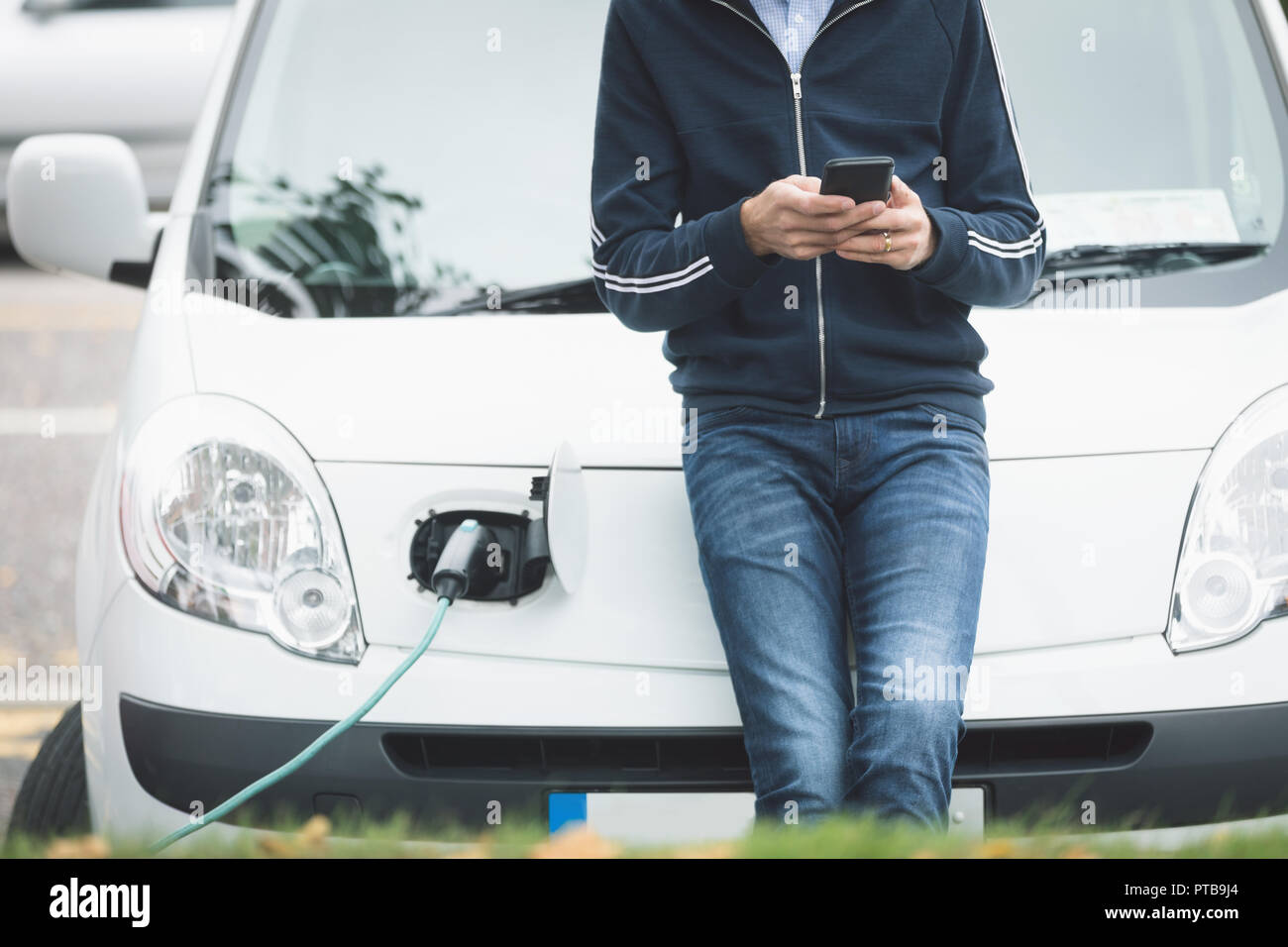 Man using mobile phone while charging electric car at charging station ...
