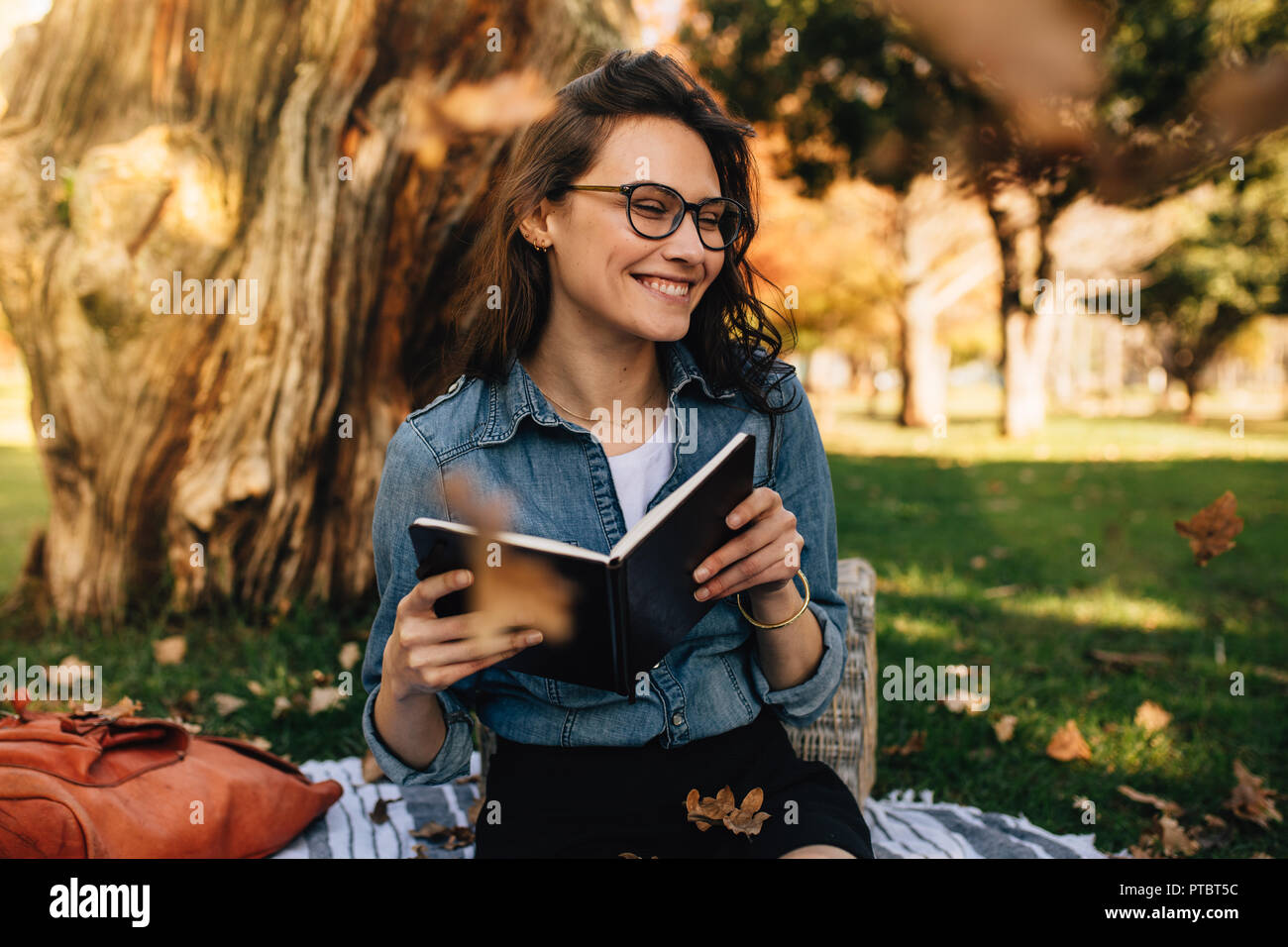 Smiling woman sitting at park holding a book with leaves falling around ...