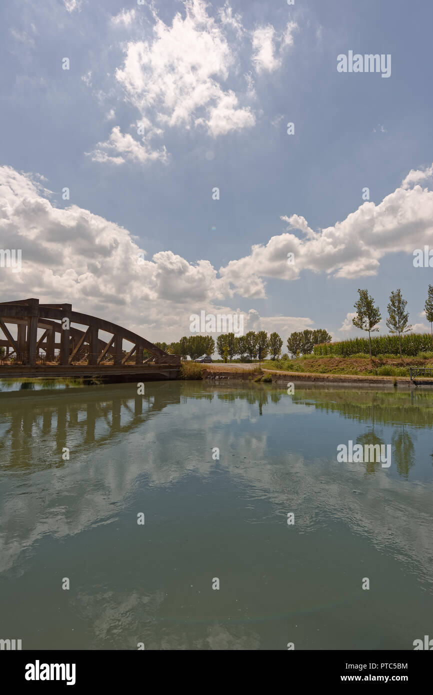 Mirror reflections on the river with bridge, clouds and row of trees ...