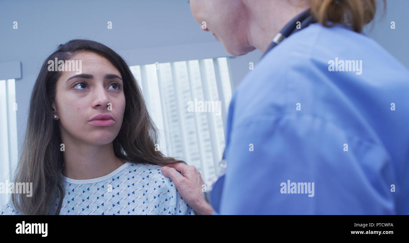 Mid aged nurse comforts young latina patient wearing hospital gown ...