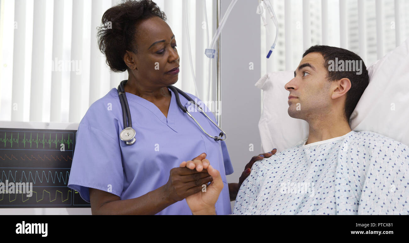 Senior black nurse comforting young hispanic patient on hospital bed ...