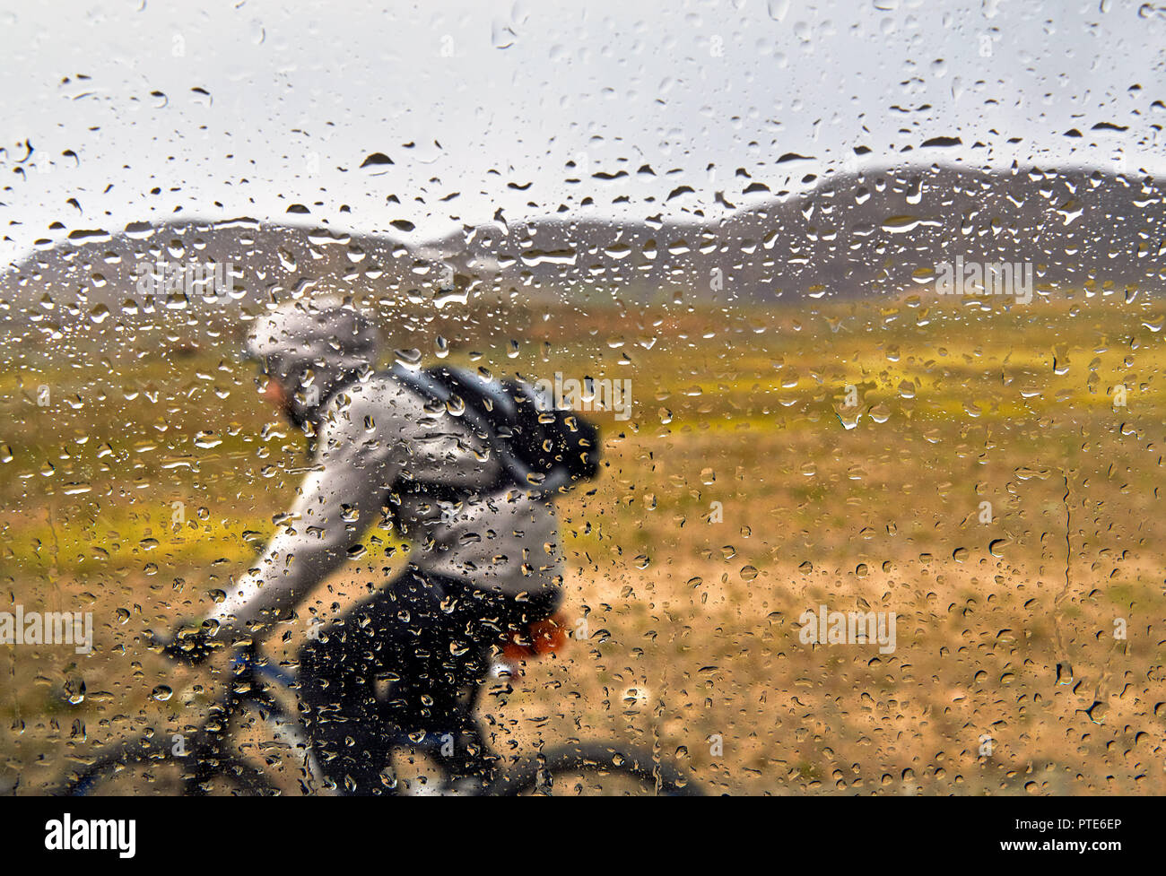 Window in rain drops and man on mountain bike rides on the country road ...
