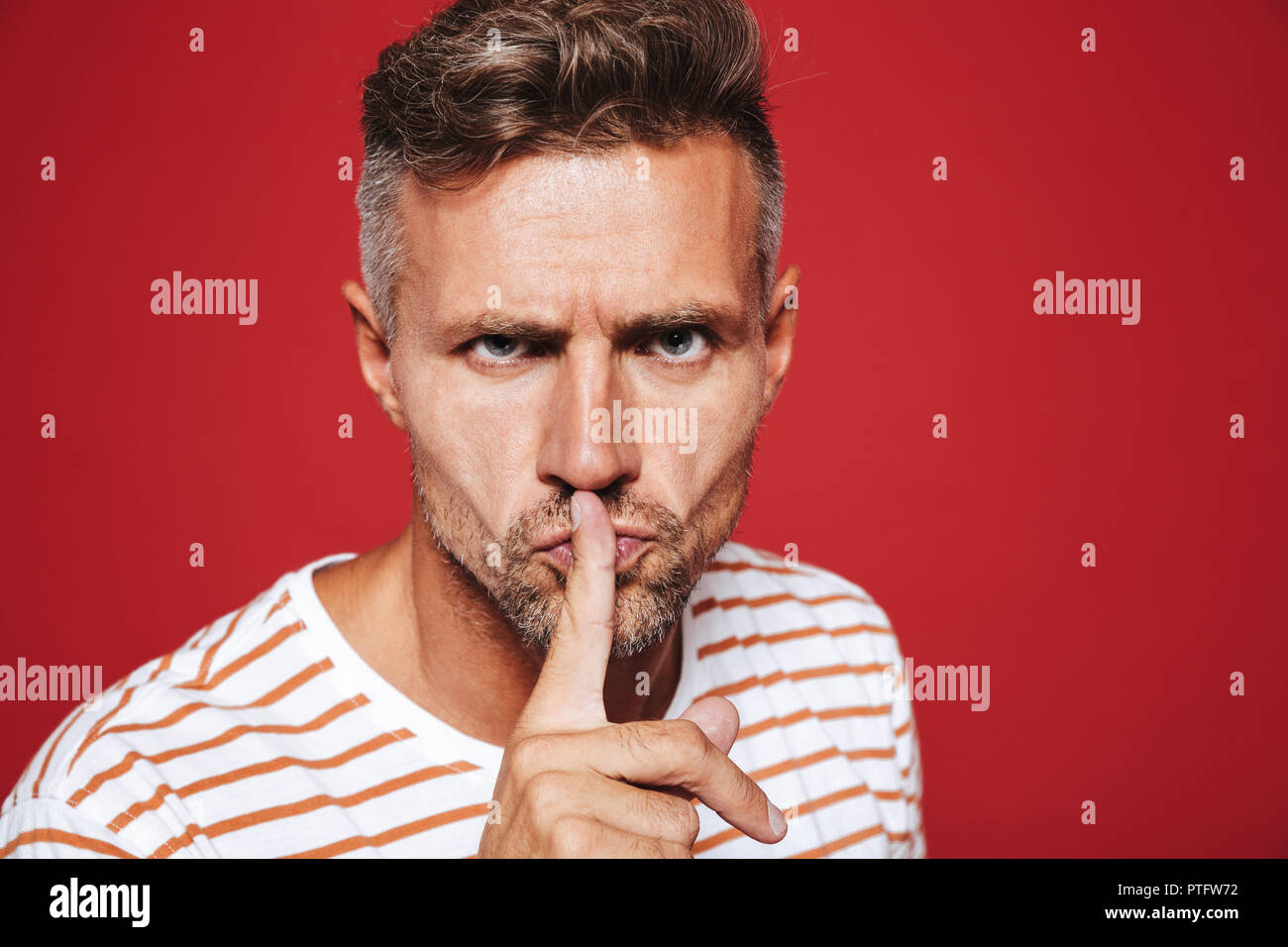 Serious man in striped t-shirt holding index finger on lips with strict ...