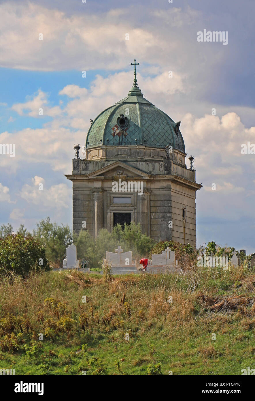 Old faded cemetery chapel building with worker tidying up grave stones ...