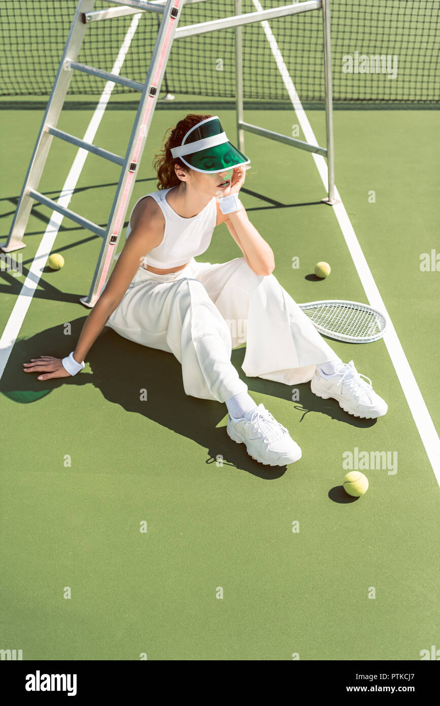 young woman in fashionable white clothing and cap sitting on tennis ...