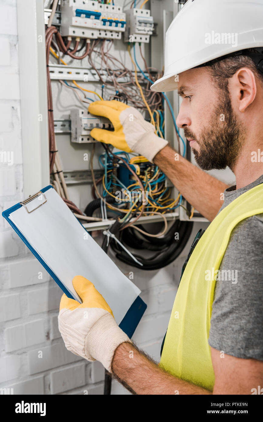 handsome electrician holding clipboard and checking electrical panel in ...
