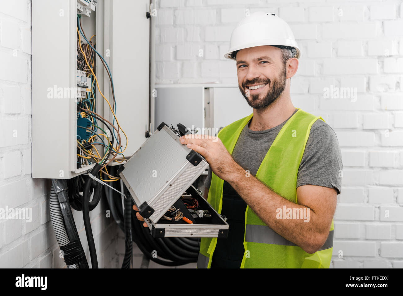 smiling electrician holding toolbox near electrical box in corridor and ...