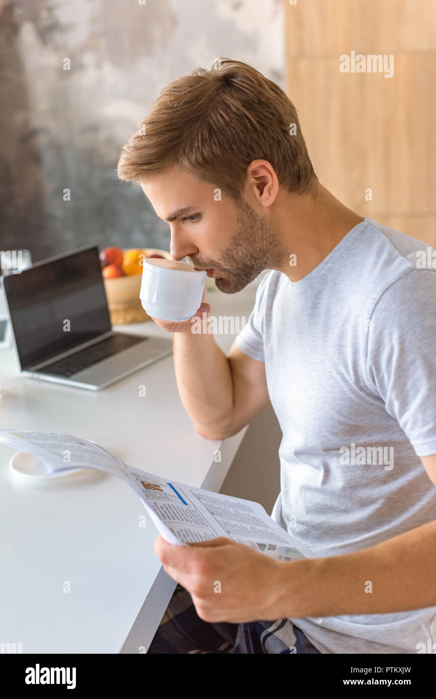 concentrated young man drinking coffee and reading newspaper at kitchen ...